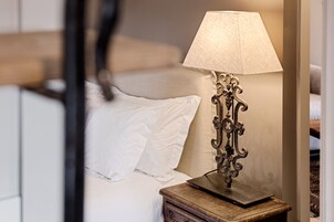 Cama bem arrumada com lençóis brancos, mesa de cabeceira de madeira com abajur e espelho na parede.