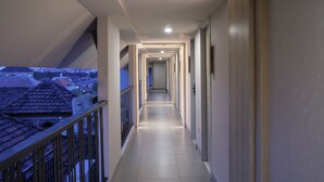 Corredor comprido e estreito com uma varanda de um lado e portas do outro, dando acesso a um quarto com vista para edifícios.