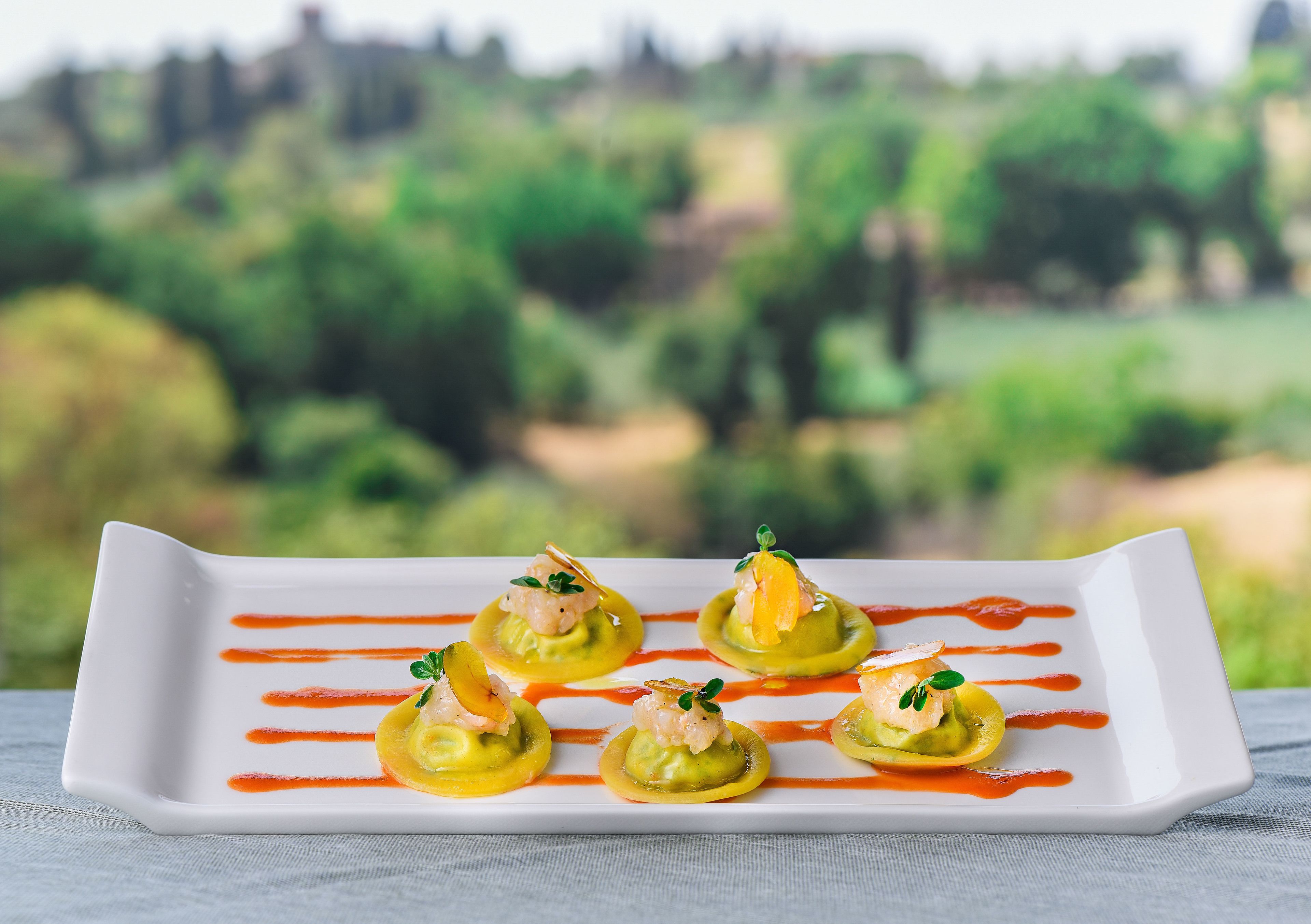 Prato branco retangular com cinco pratos de ravioli, cada um decorado com um ervo verde e uma fatia de fruta laranja, dispostos sobre uma mesa com uma paisagem verde desfocada ao fundo.