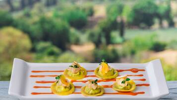 Prato branco retangular com cinco pratos de ravioli, cada um decorado com um ervo verde e uma fatia de fruta laranja, dispostos sobre uma mesa com uma paisagem verde desfocada ao fundo.