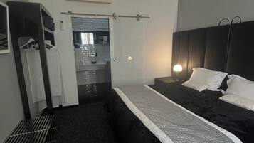 Quarto de hotel com cama grande, mesa de cabeceira, armário e banheiro visível por uma porta aberta.