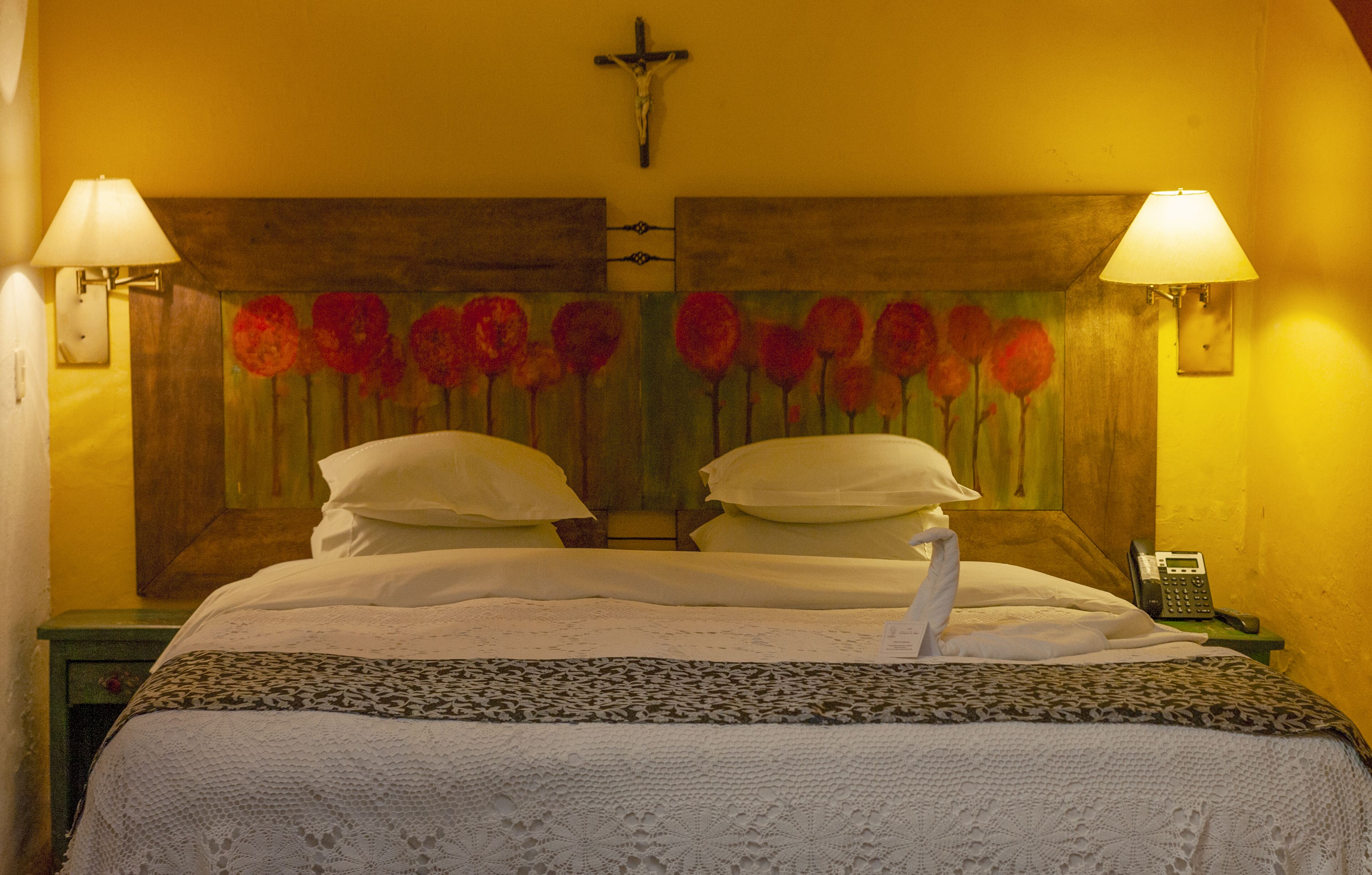 Quarto de hotel com cama, duas luminárias fixadas na parede, cabeceira de madeira com arte floral, uma cruz na parede e telefone no criado-mudo.