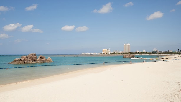 Uma praia com águas turquesa cristalinas, um afloramento rochoso e edifícios ao fundo.