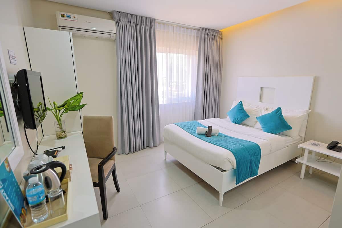Quarto de hotel com cama, mesa de cabeceira, cadeira, escrivaninha com um computador e janela com cortinas.