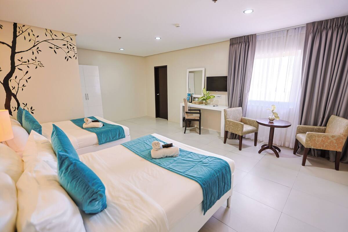 Quarto de hotel com duas camas, uma escrivaninha, uma cadeira, uma televisão e um guarda-roupa.