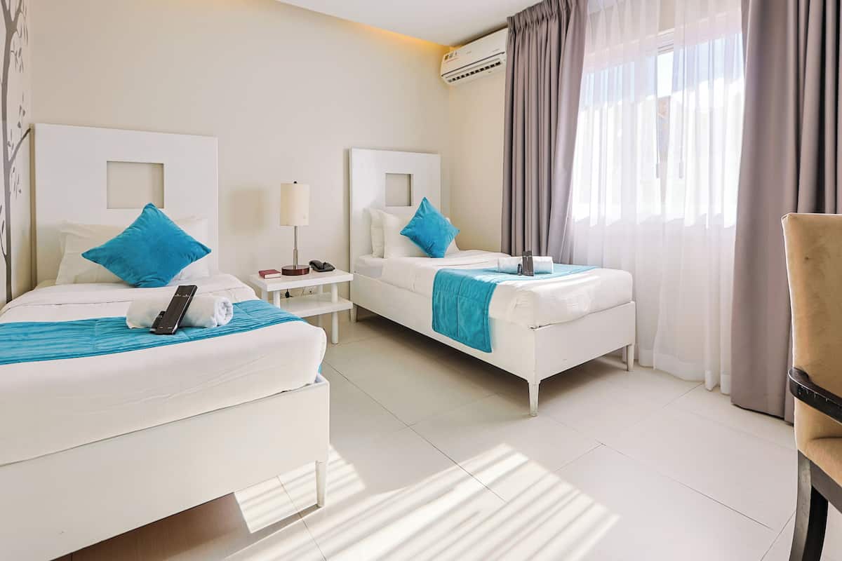 Quarto de hotel com duas camas, mesa de cabeceira, cadeira e janela com cortinas.