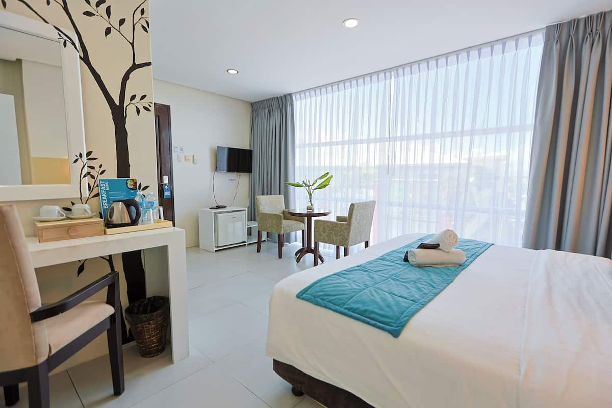 Quarto de hotel com cama, escrivaninha com cadeira, mesa pequena com cadeiras, televisão e janela com cortinas.
