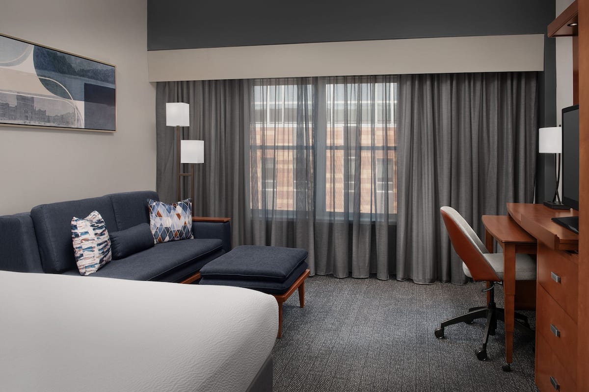 Quarto de hotel com cama, sofá, escrivaninha e uma janela grande com cortinas.