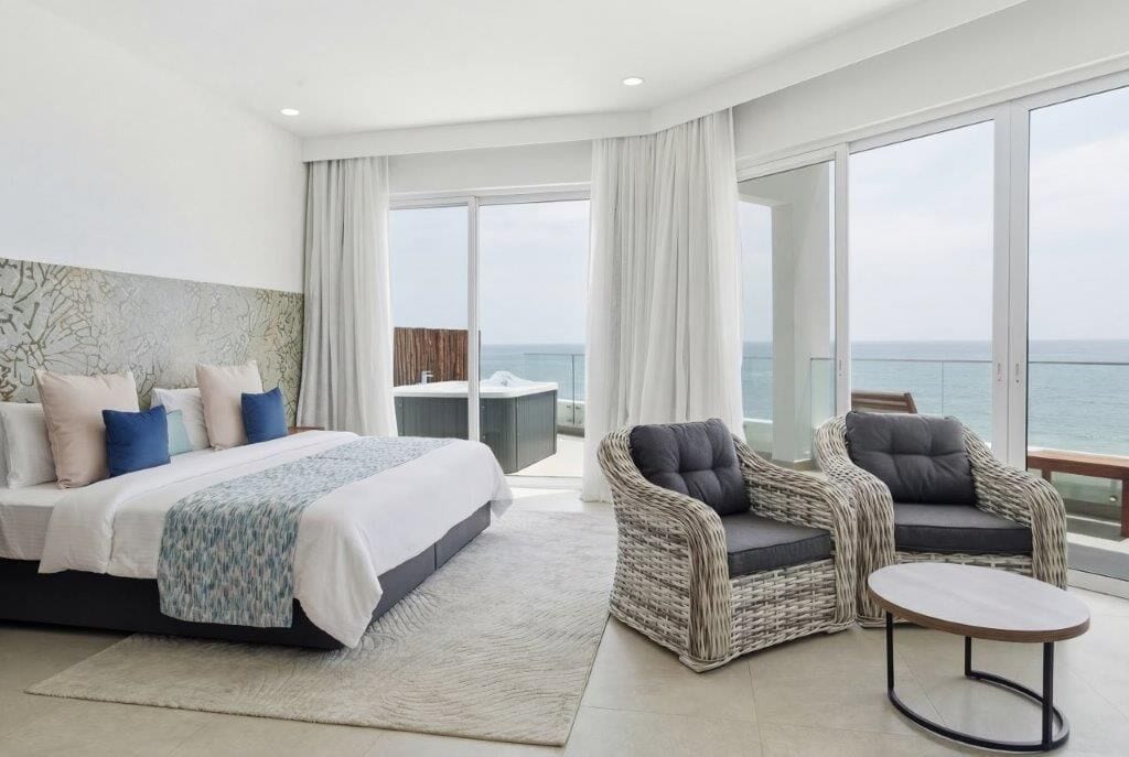 Quarto com cama, duas cadeiras de vime, uma mesa pequena e vista para o mar.