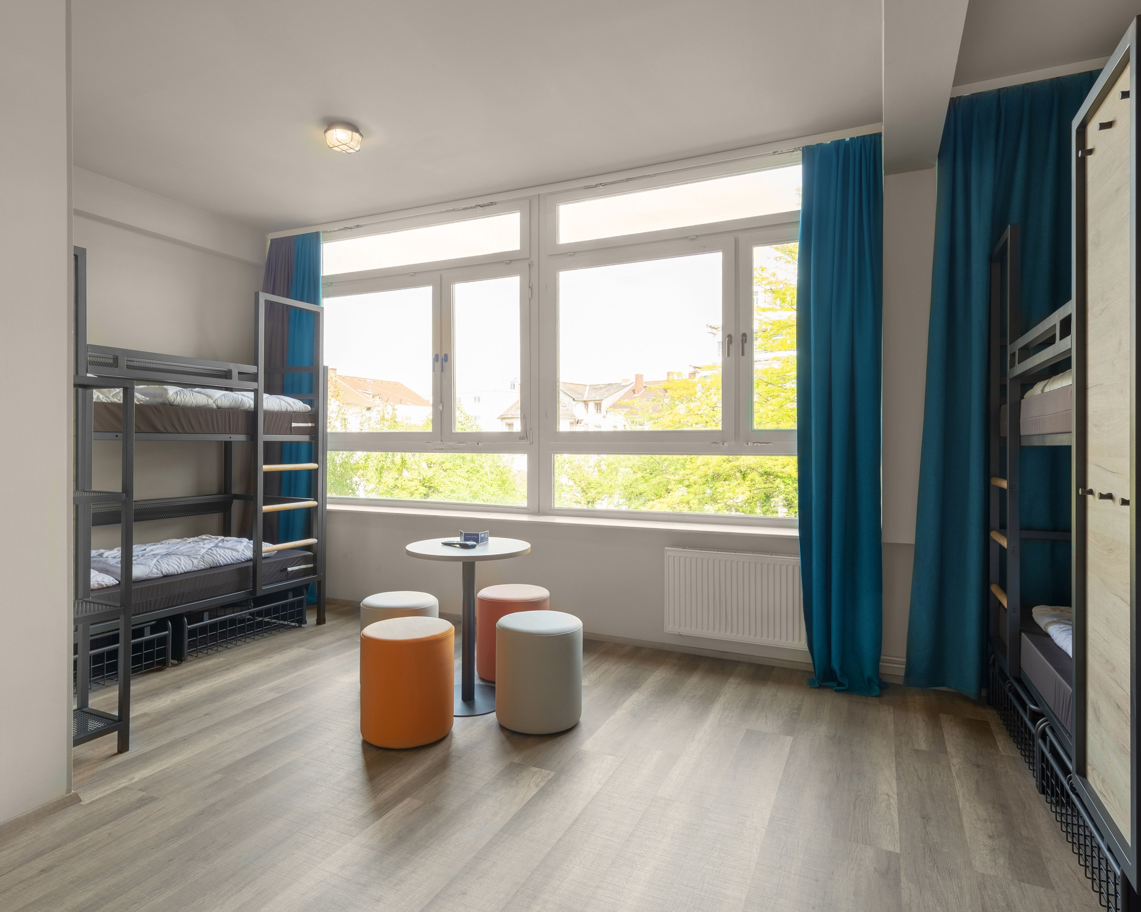 Quarto em dormitório com beliches, uma pequena mesa redonda, dois banquinhos e uma janela com cortinas.