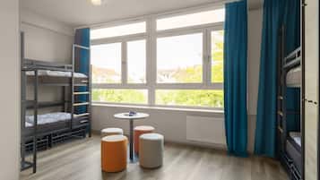 Quarto em dormitório com beliches, uma pequena mesa redonda, dois banquinhos e uma janela com cortinas.