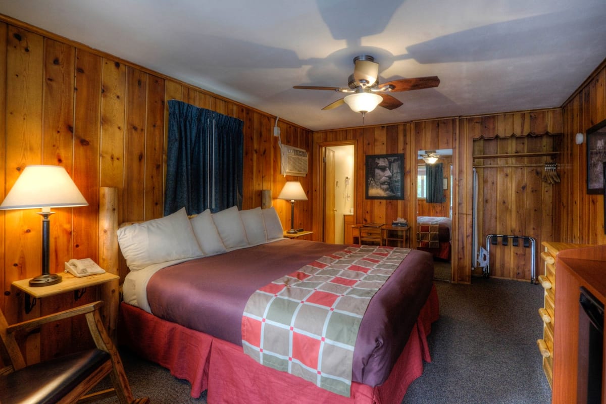 Um quarto com painéis de madeira, uma cama grande, duas mesas de cabeceira com luminárias, um ventilador de teto e um armário de madeira.