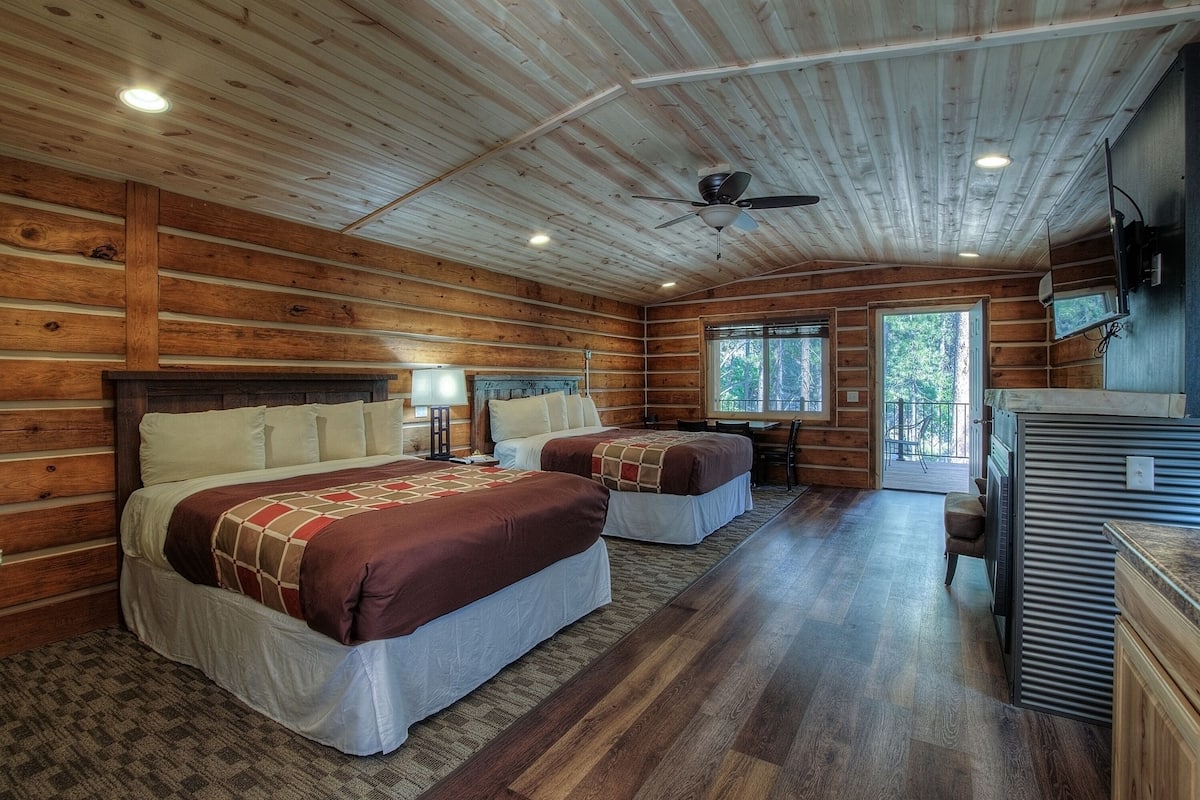 Quarto de cabana de madeira com duas camas, um ventilador de teto, uma TV e janela com vista para a floresta.