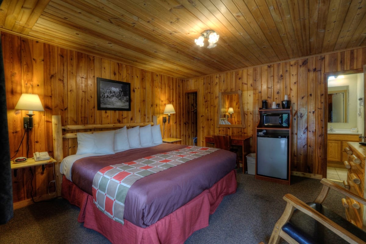 Quarto de cabana de madeira com uma cama, duas luminárias de cabeceira, uma escrivaninha pequena com cadeira, um refrigerador e um banco de madeira.