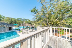 Um deck de madeira com vista para uma piscina, com cadeiras de praia e guarda-sóis, cercado por árvores e um edifício distante.
