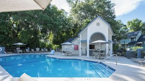Piscina com espreguiçadeiras e guarda-sóis, cercada por árvores e um edifício com varanda coberta.
