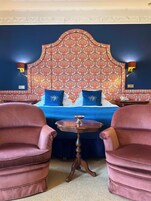 Quarto de hotel com uma cama grande, duas almofadas azuis, cabeceira decorativa, duas poltronas, uma mesinha e luminárias na parede.