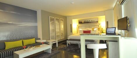 Quarto de hotel moderno com uma cama grande, uma escrivaninha com computador, um sofá com almofadas e um quadro na parede.