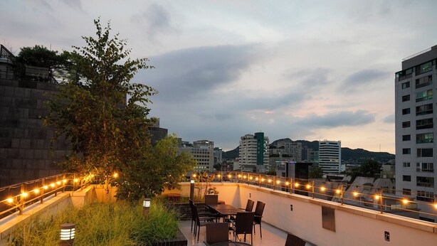 Área de estar ao ar livre com mesas e cadeiras, rodeada por gramíneas altas e uma paisagem urbana ao fundo.