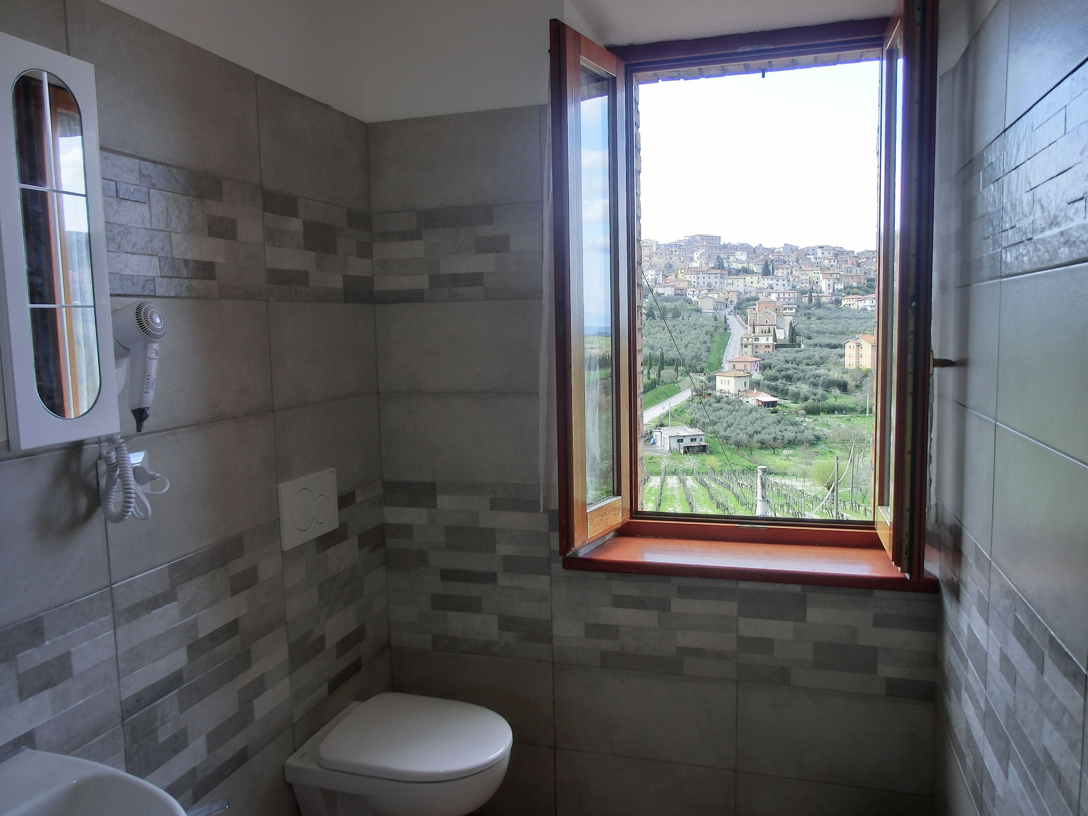 Casa de banho com sanita, secador de cabelo e janela com vista para uma vila.