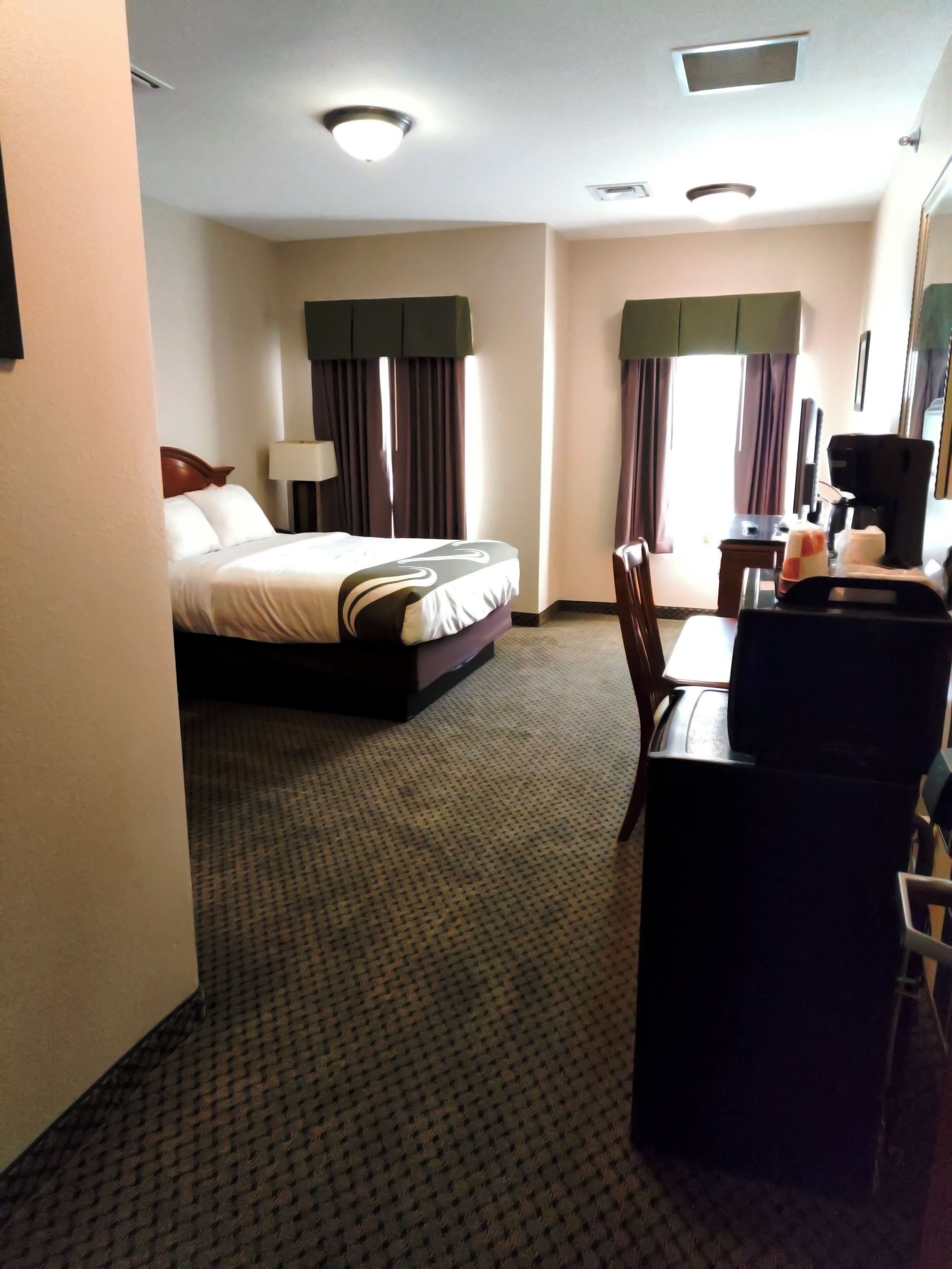 Quarto de hotel com cama, escrivaninha, cadeira, televisão e janela com cortinas.