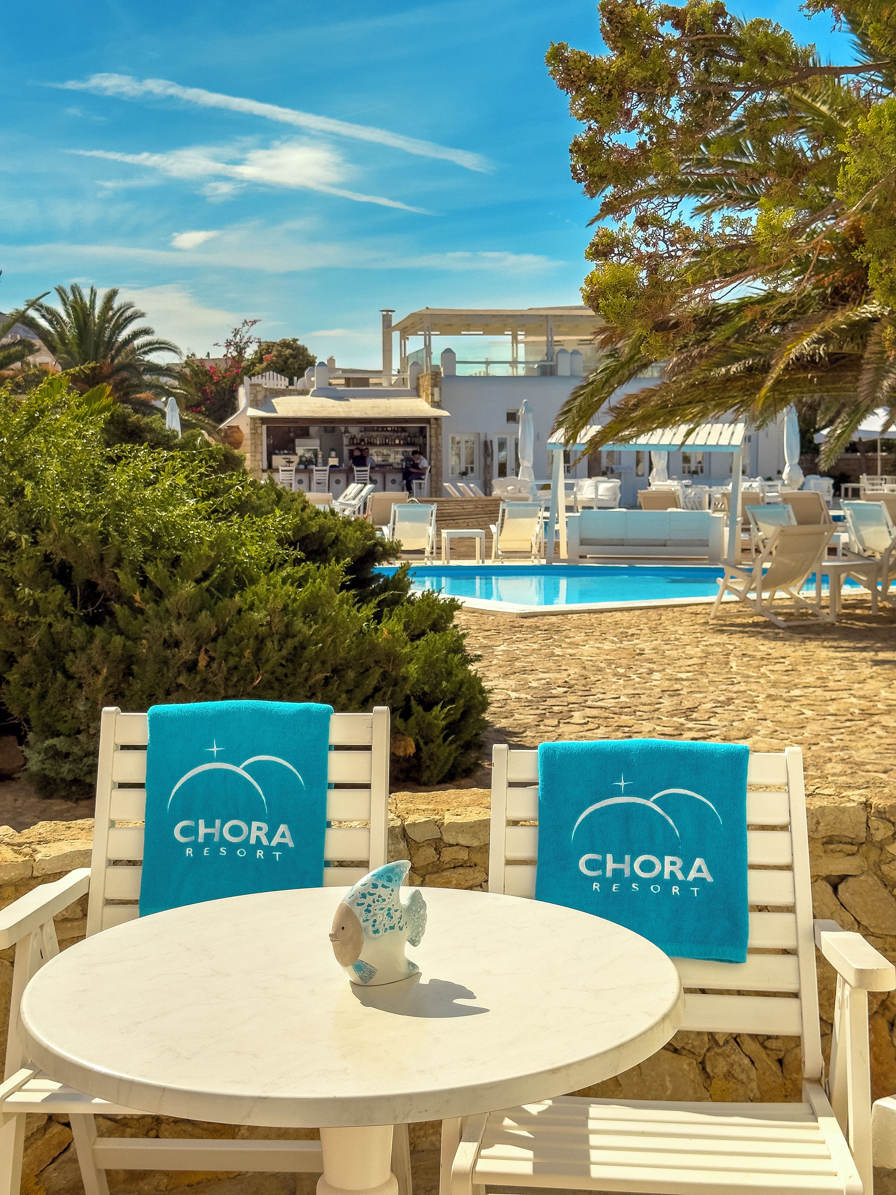 Área da piscina com espreguiçadeiras, mesa e vista para um edifício com bar.