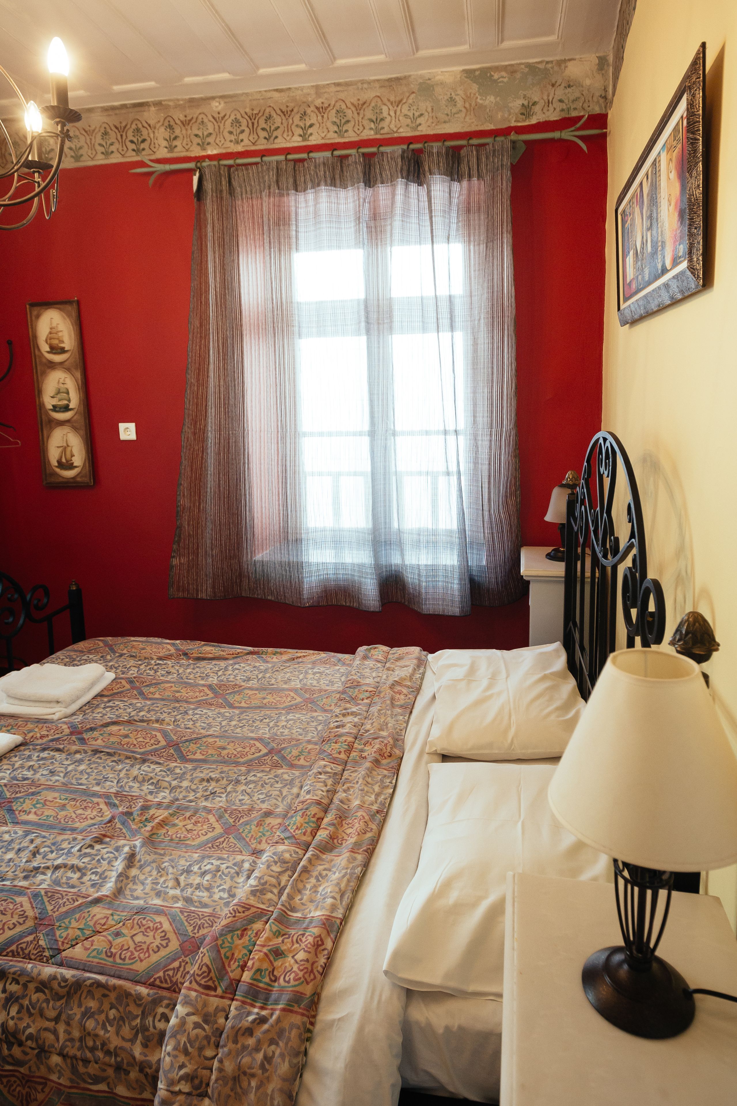 Quarto com colcha estampada, mesa de cabeceira com abajur, parede vermelha e janela com cortinas leves.