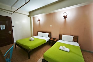 Quarto de hotel com duas camas de solteiro, cada uma com colcha verde e travesseiros brancos, mesa de cabeceira de madeira com telefone e porta marrom.