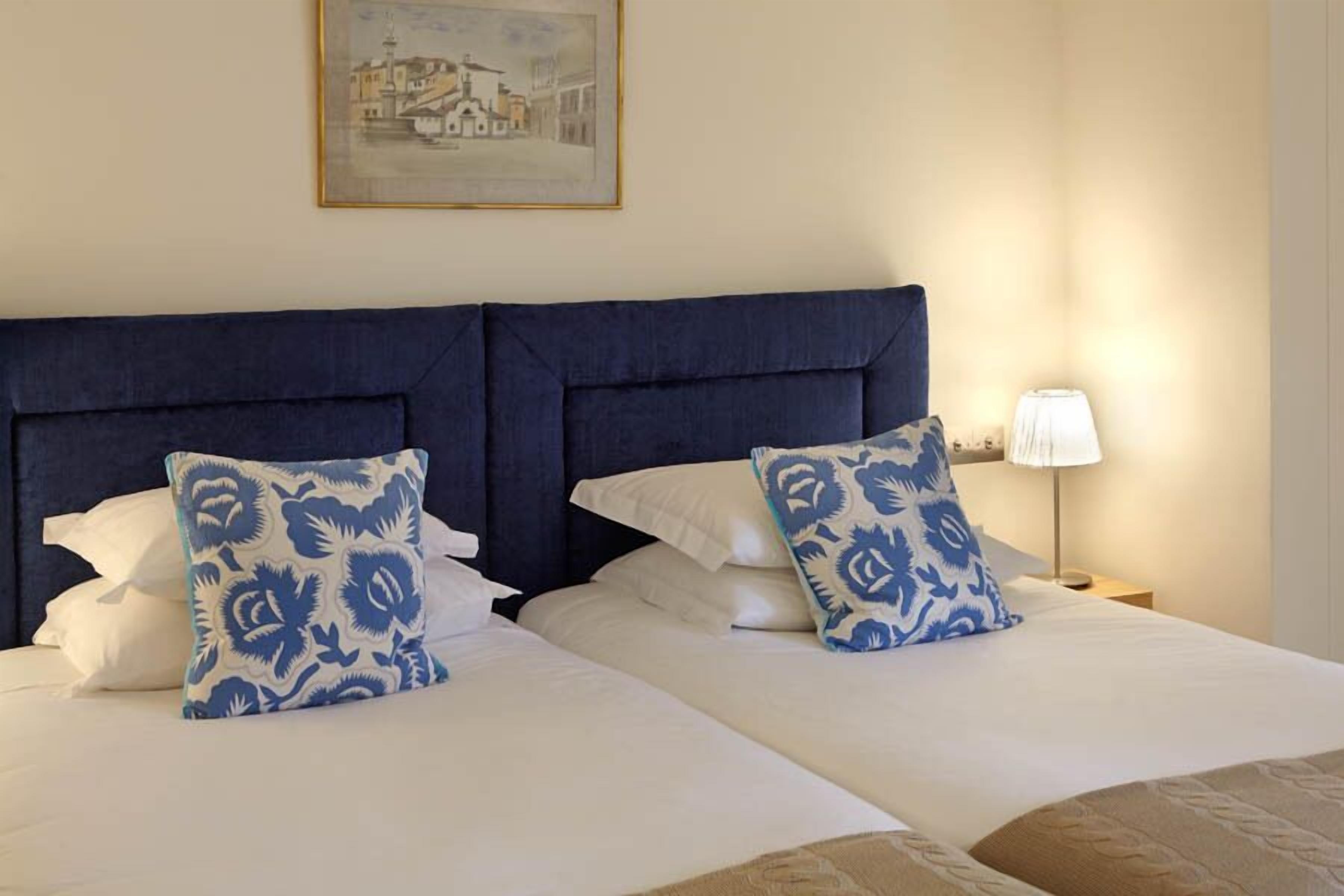 Cama de casal com cabeceira escura, roupa de cama branca e duas almofadas azuis estampadas. É visível uma mesa de cabeceira com um abajur.