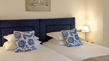 Cama de casal com cabeceira escura, roupa de cama branca e duas almofadas azuis estampadas. É visível uma mesa de cabeceira com um abajur.