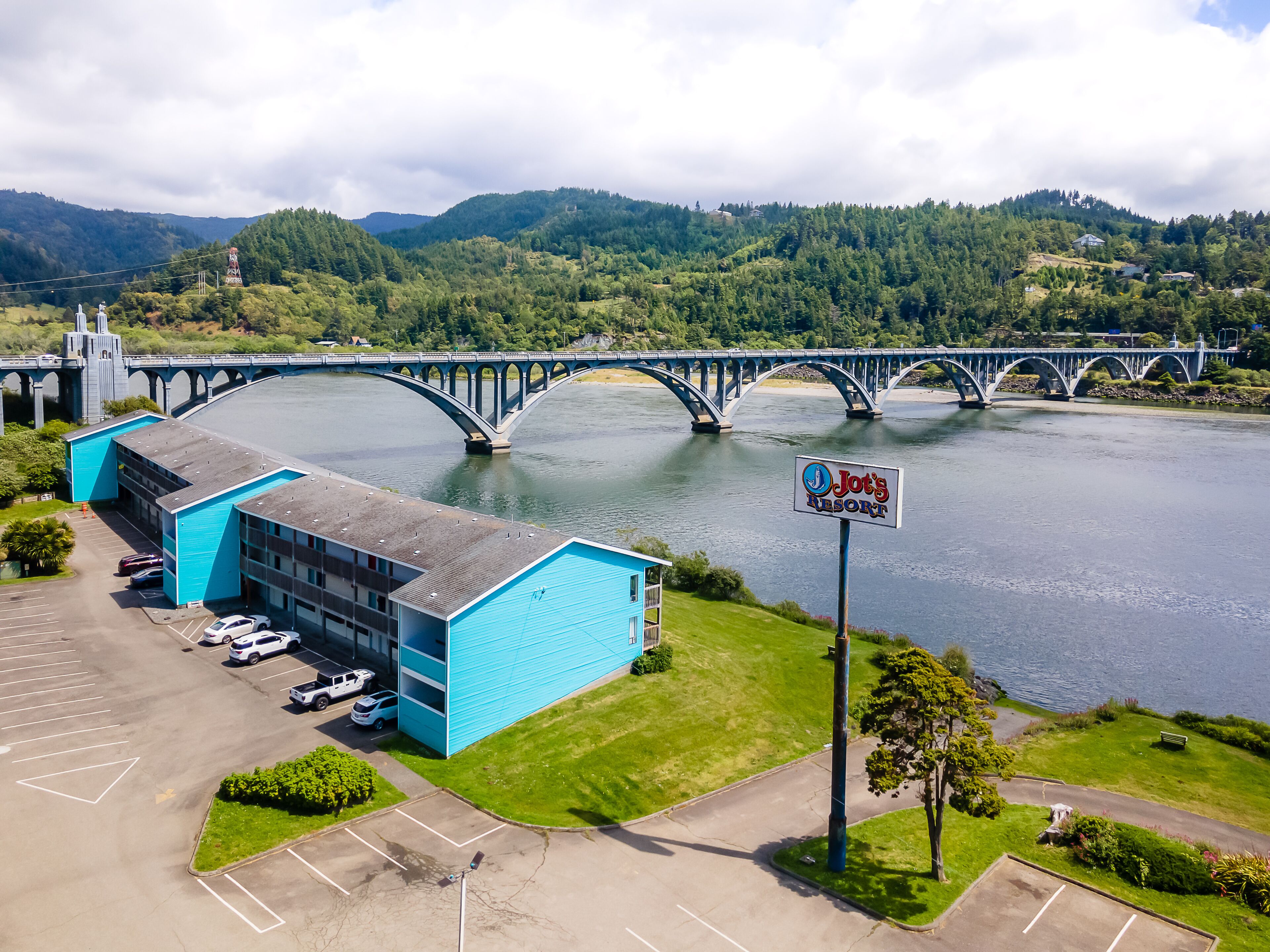 Resort com um edifício azul, estacionamento e uma ponte sobre o rio.