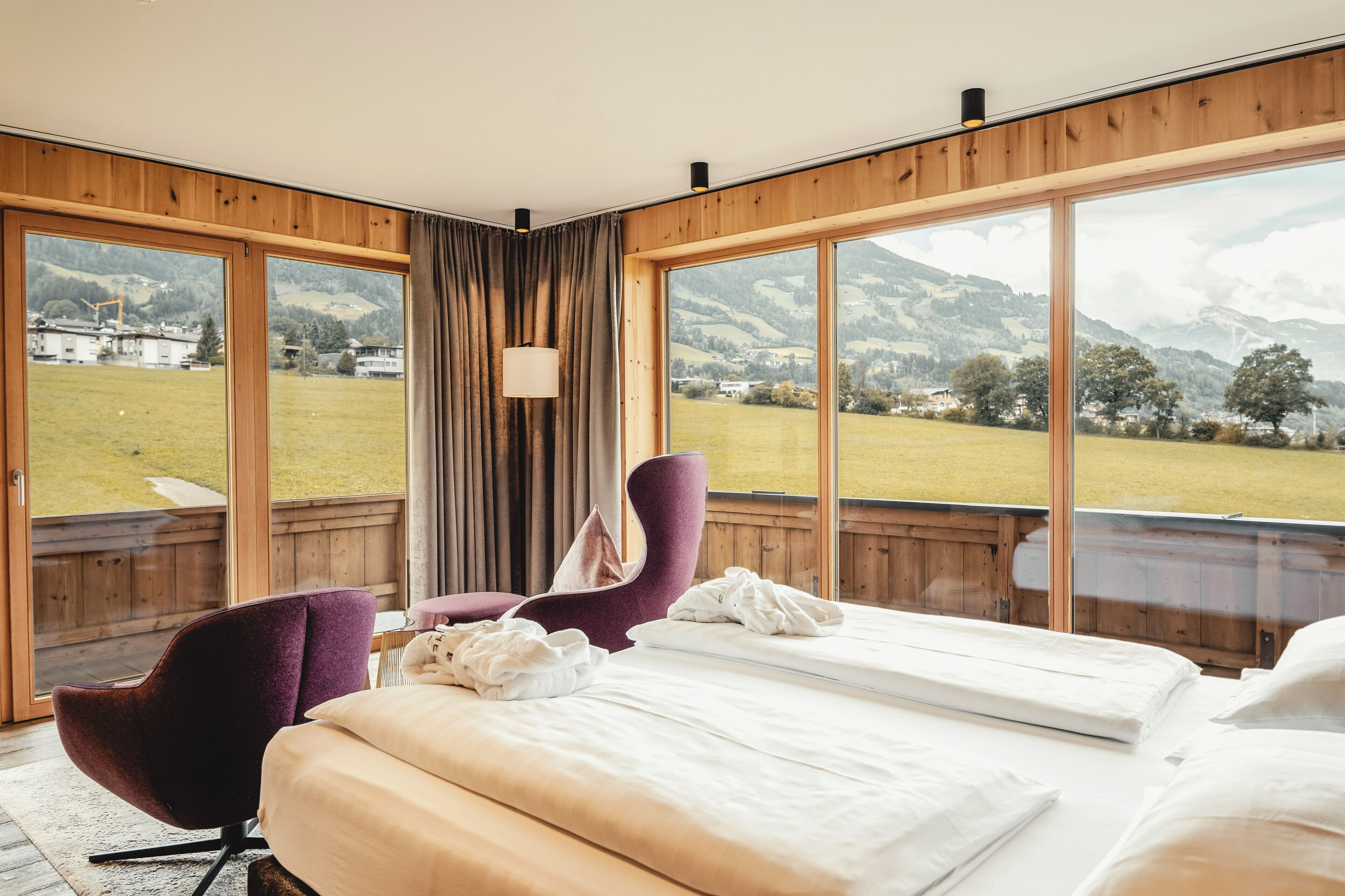 Quarto com uma cama grande, uma poltrona roxa e vista para uma paisagem montanhosa através de grandes janelas.