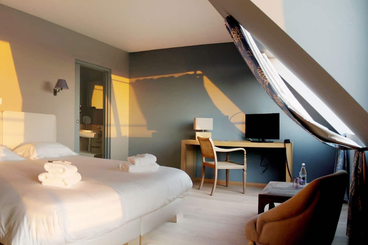 Quarto de hotel com cama, escrivaninha, cadeira, televisão e janela com cortinas.