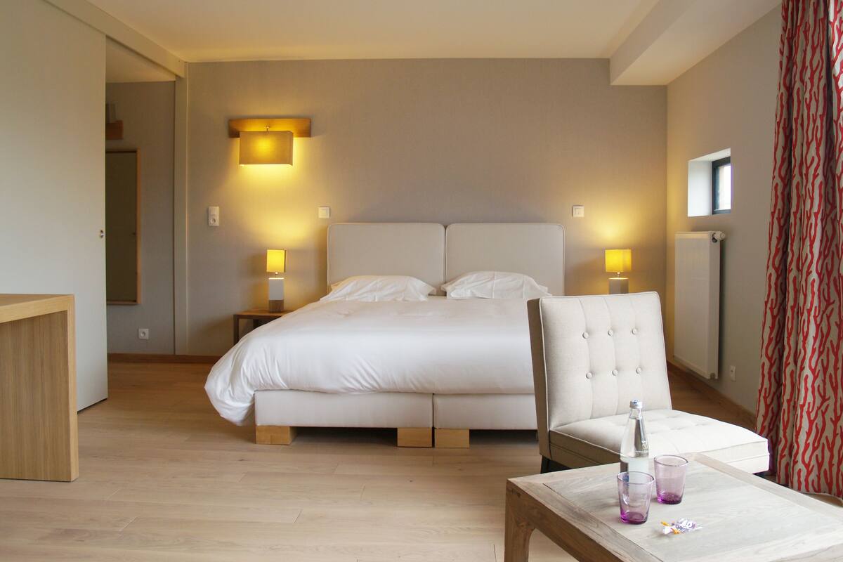 Quarto de hotel com uma cama grande, duas mesas de cabeceira com luminárias, um criado-mudo de madeira, uma mesinha com copos e uma garrafa, e cortina vermelha estampada.