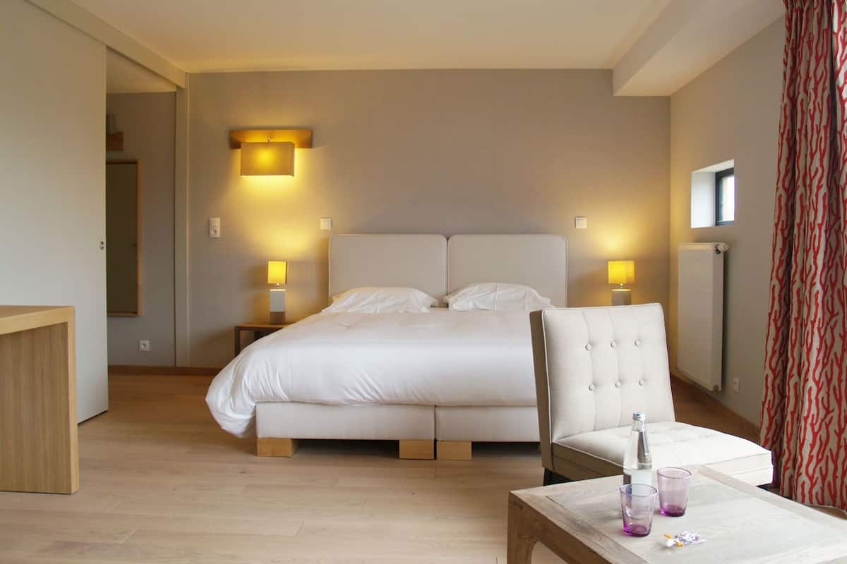 Quarto de hotel com uma cama grande, abajures, uma mesa de cabeceira de madeira, uma pequena mesa com copos e uma cortina vermelha com padrão.