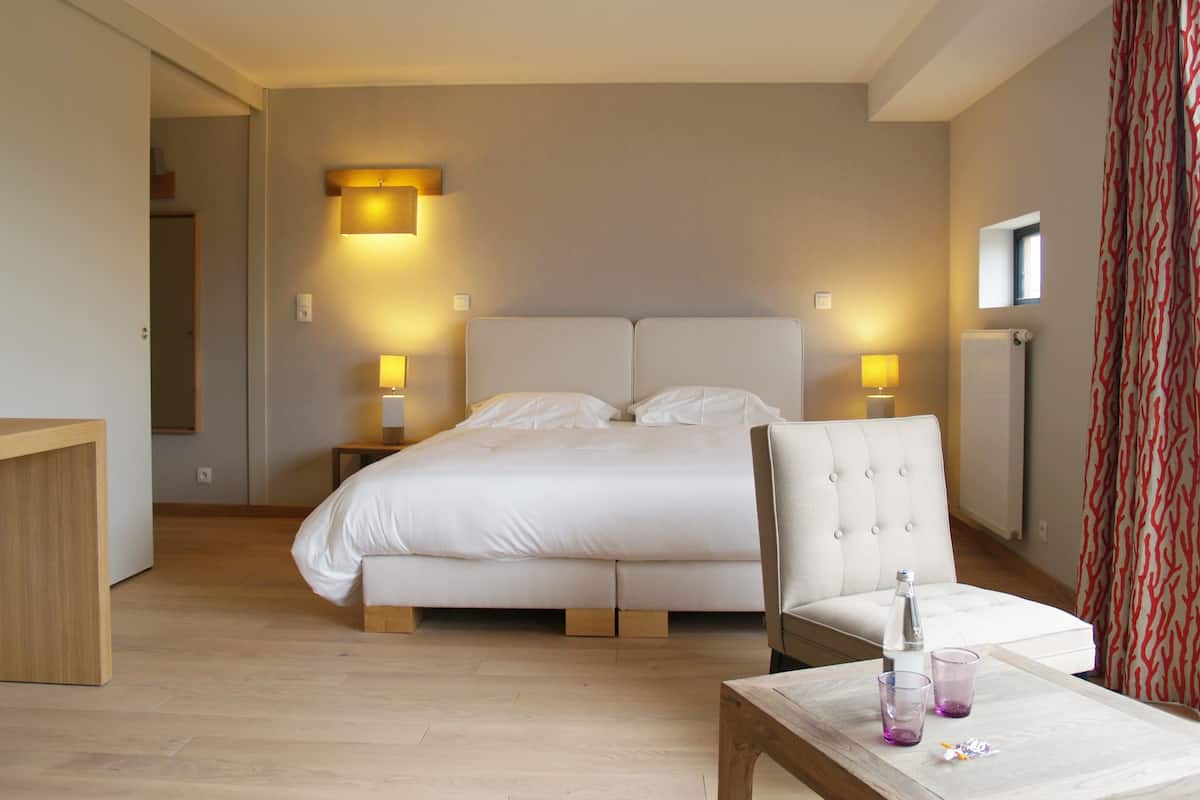 Quarto de hotel com cama, abajures, mesa de cabeceira de madeira, mesa com copos e cortina estampada.