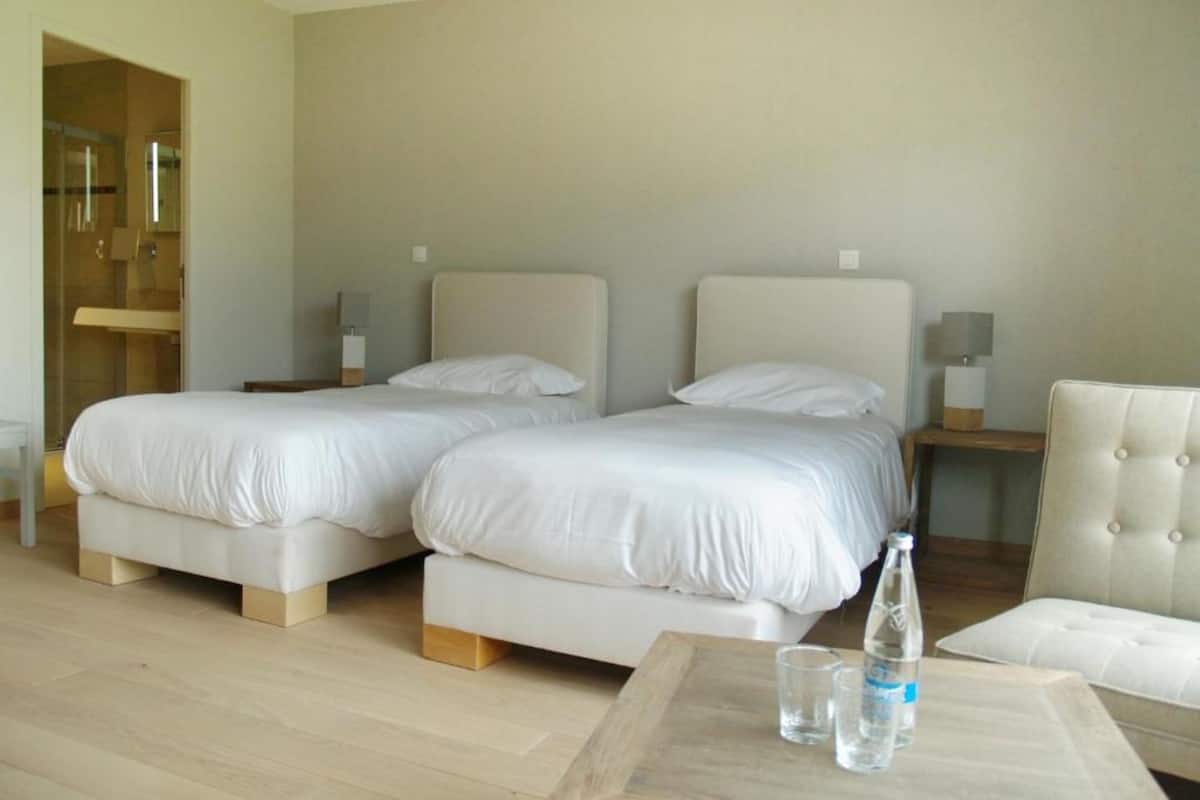 Quarto de hotel com duas camas, piso de madeira, uma cadeira e uma mesa com garrafa e copos.