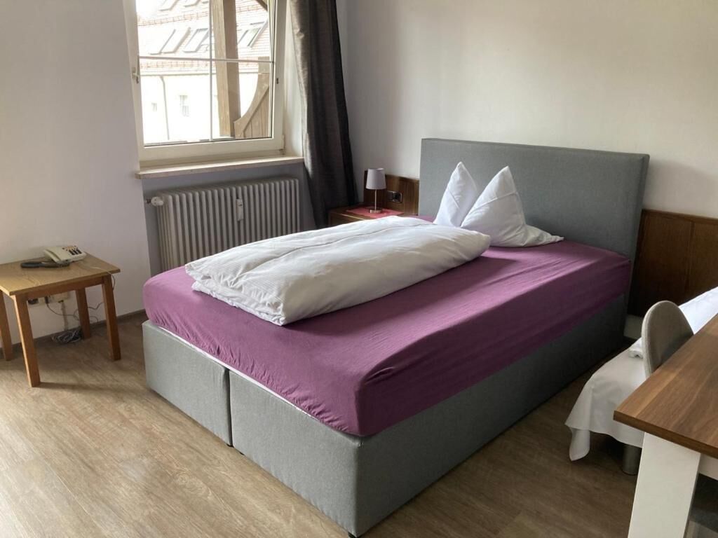 Cama bem arrumada com cabeceira cinza e colcha roxa, uma pequena mesa de cabeceira com abajur, e uma janela com cortinas.