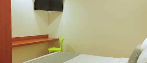 Quarto de hotel com cama, televisão na parede, uma mesinha e uma cadeira verde.