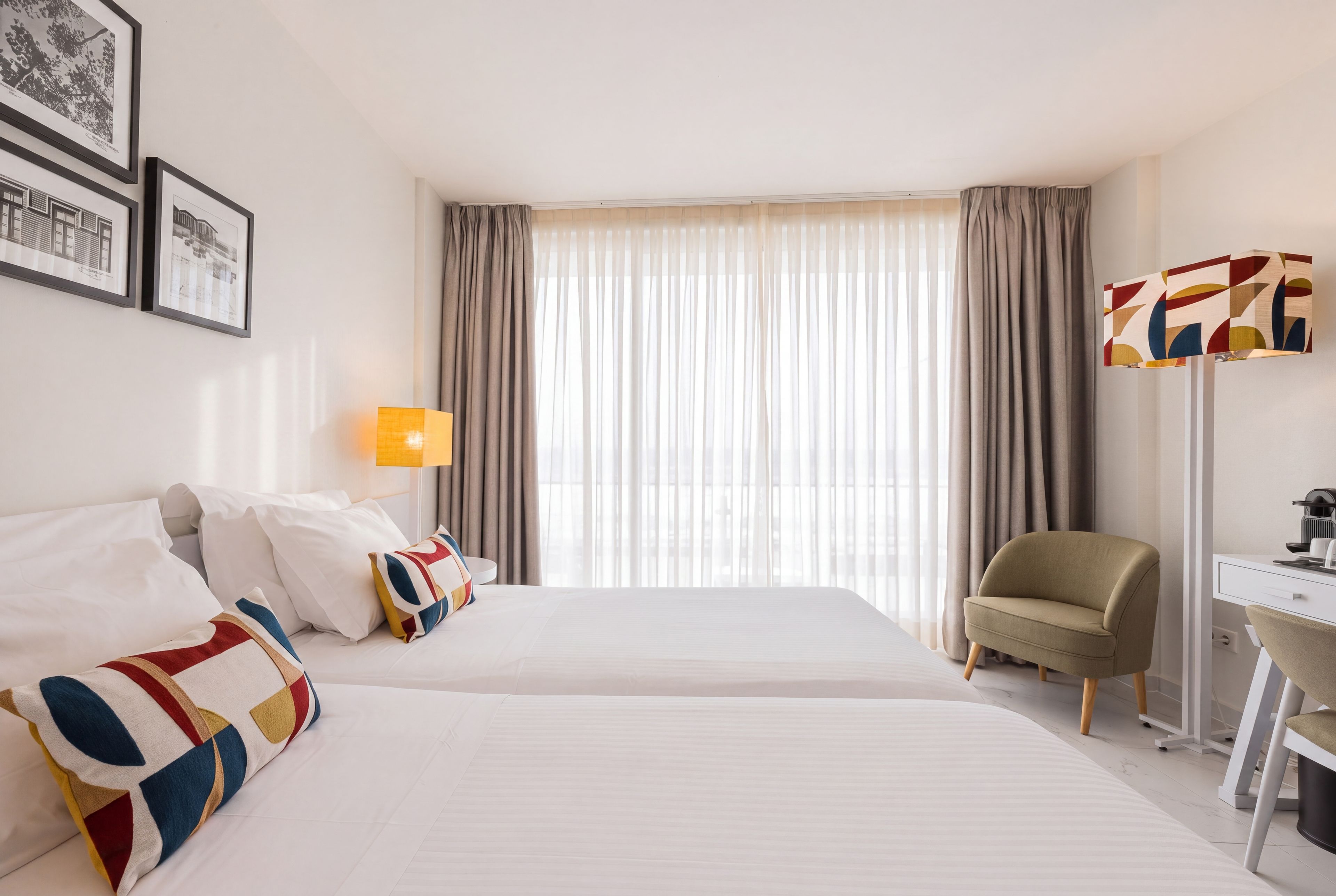 Quarto de hotel com duas camas, uma escrivaninha, uma cadeira, um abajur e cortinas.