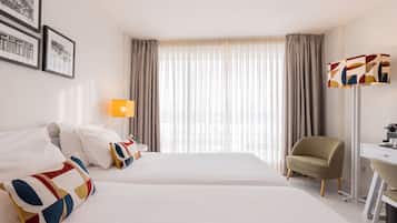 Quarto de hotel com duas camas, uma escrivaninha, uma cadeira, um abajur e cortinas.