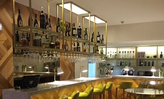 Uma área de bar com prateleiras de garrafas de bebidas e copos, um balcão com um computador e cadeiras verde-claras.