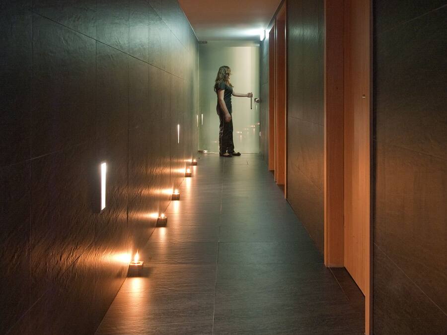Um corredor iluminado por pequenas velas no chão, com uma pessoa caminhando em direção a uma porta.