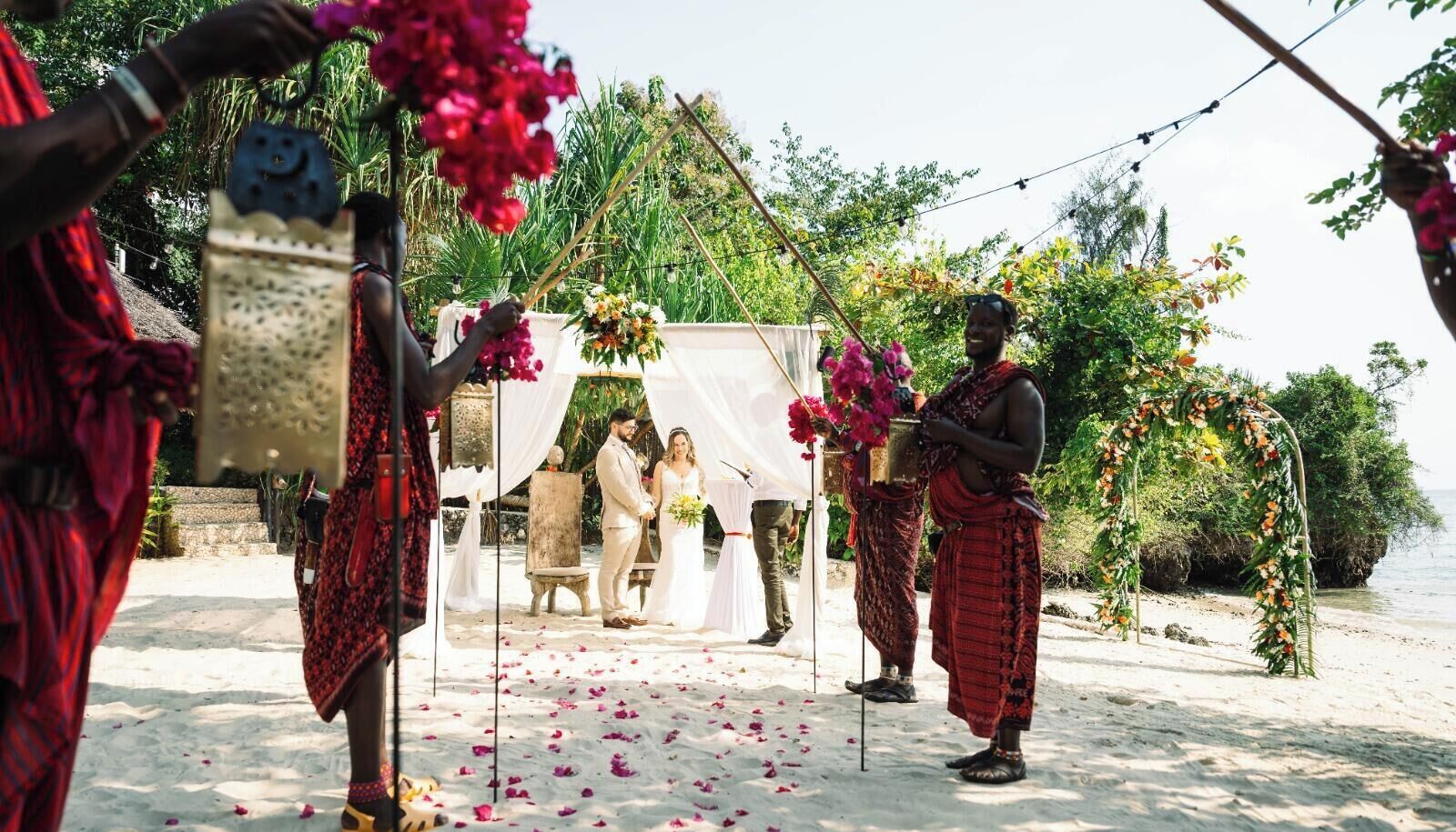 Uma cerimônia de casamento na praia, com uma tenda branca, decorações florais e convidados com trajes típicos.