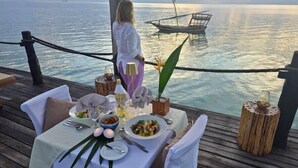 Uma mesa de jantar posta com pratos, talheres e uma garrafa, com vista para um mar calmo e um barco ao longe.