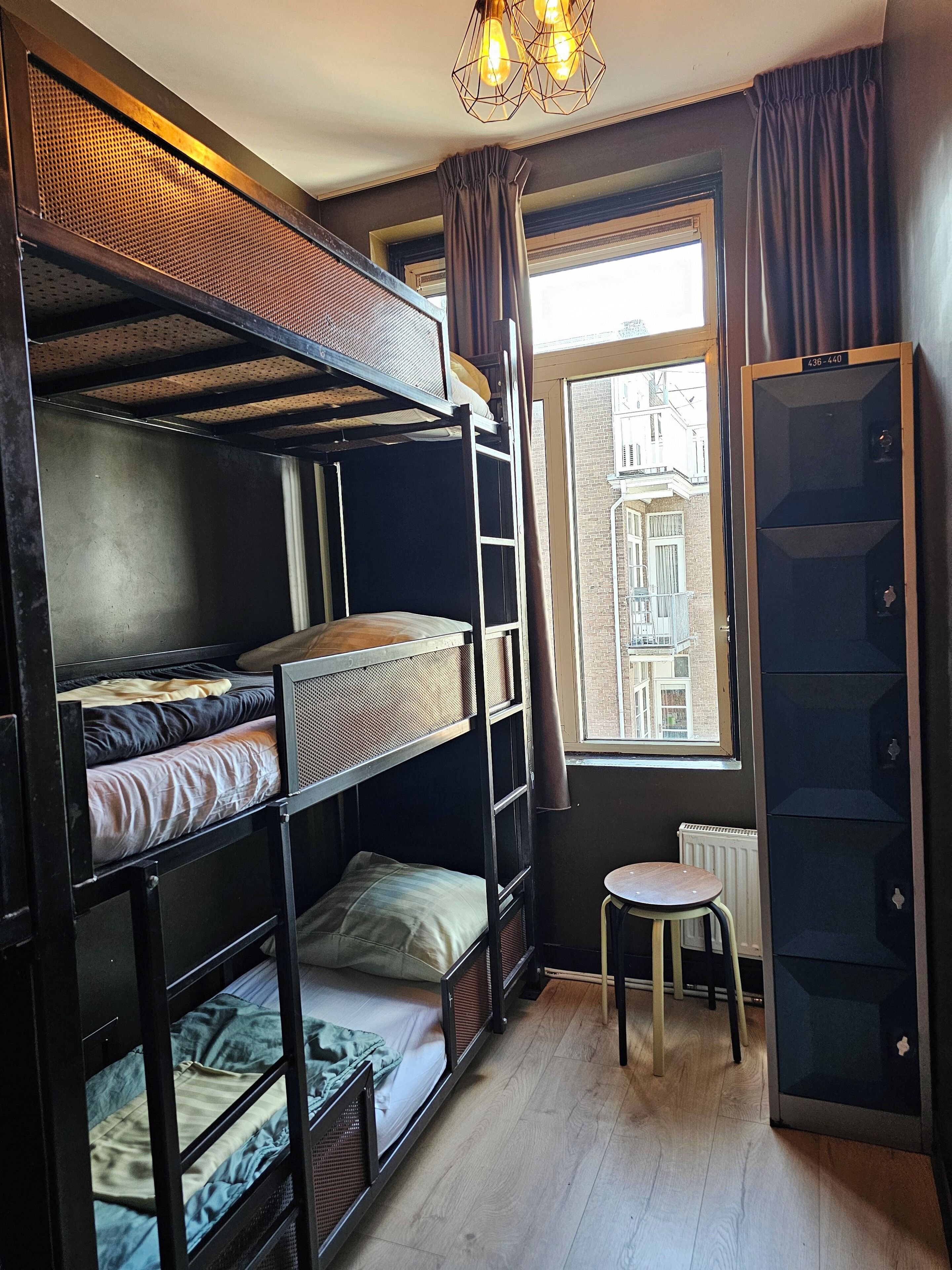 Quarto de dormitório com beliches, um radiador, uma mesinha e uma janela com vista para edifícios.