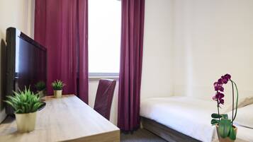 Quarto de hotel com cama, escrivaninha com duas plantas em vasos, televisão e janela com cortinas roxas.