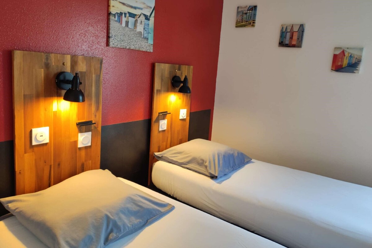 Quarto de hotel com duas camas, cabeceiras de madeira, luminárias fixadas na parede e uma parede com detalhes em vermelho.