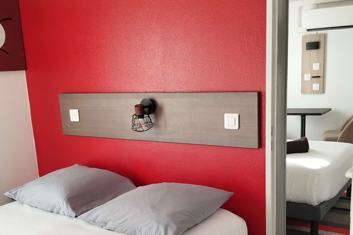 Quarto de hotel com parede de destaque vermelha, cama com roupa de cama branca e luminária moderna sobre a cama.