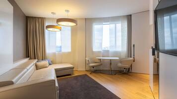 Uma sala de estar moderna com sofá, uma mesinha e duas cadeiras. Há grandes janelas com cortinas, um lustre e uma televisão de tela plana na parede.